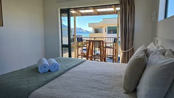 Quarto de hotel com cama, duas toalhas enroladas, varanda com mesa e cadeiras, e vista para edifícios e montanhas.