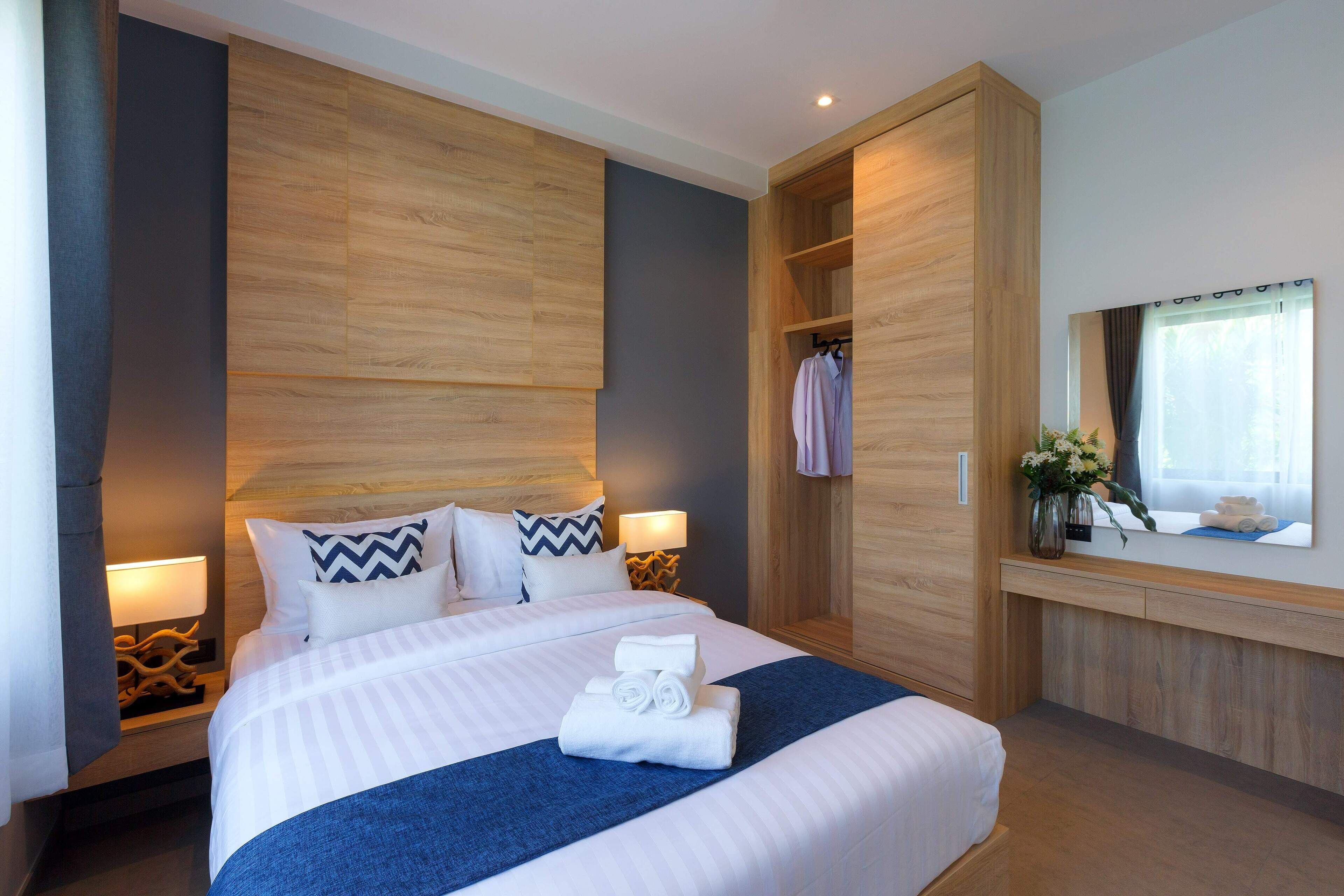 Quarto de hotel com uma cama grande, cabeceira de madeira, guarda-roupa, espelho e mesa de cabeceira com abajur.