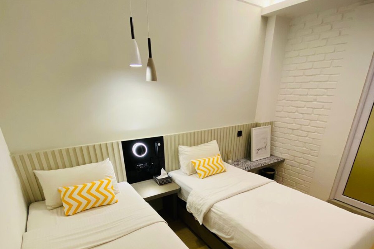 Quarto de hotel com duas camas, uma mesa de cabeceira com telefone e abajur, e parede com padrão de tijolos.
