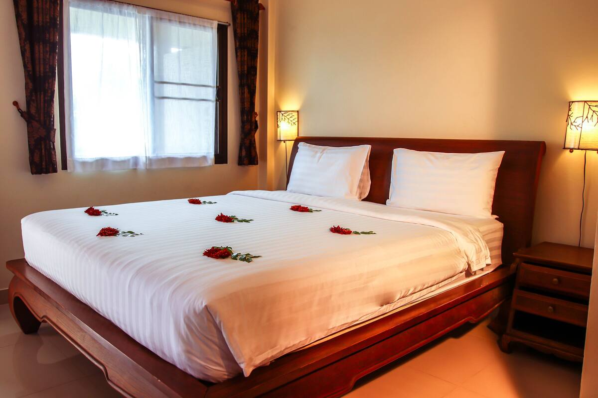 Cama bem arrumada com lençóis brancos e flores vermelhas, cabeceira de madeira e duas luminárias laterais.