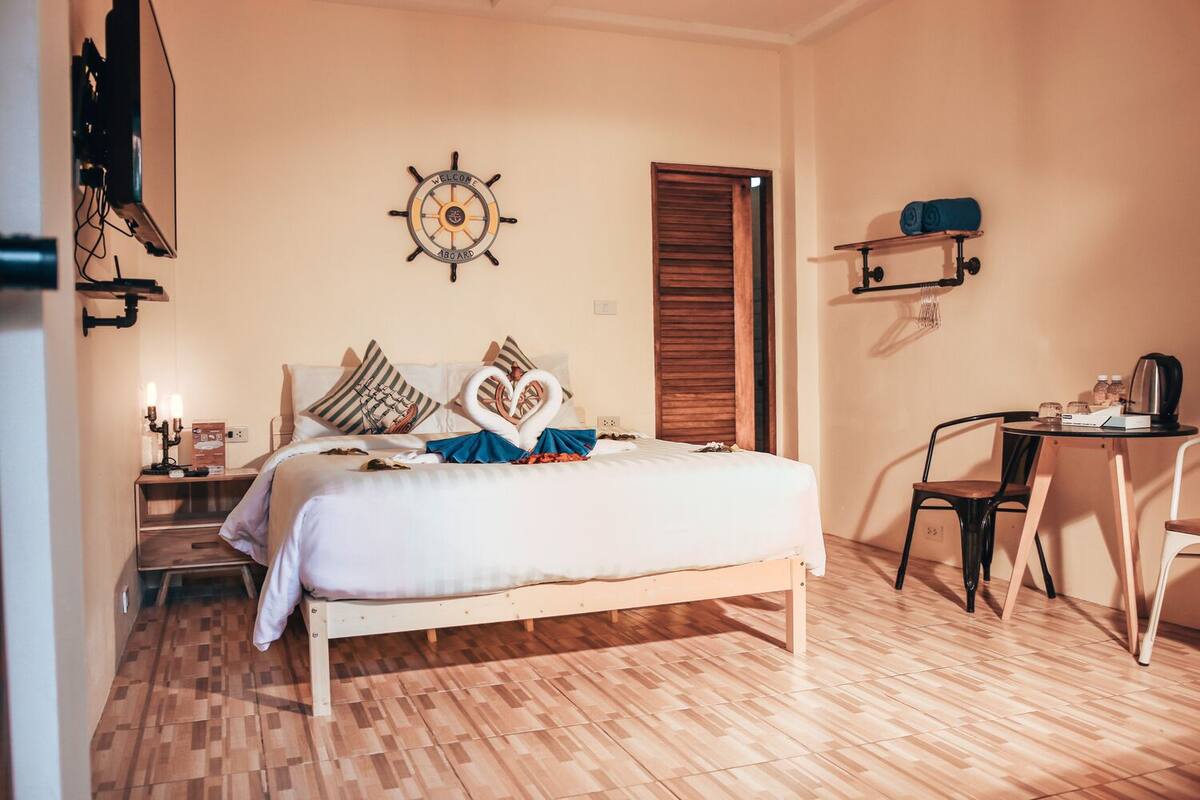 Quarto com cama, mesa de cabeceira, relógio na parede, porta de madeira, escrivaninha com cadeira e prateleira com objetos.