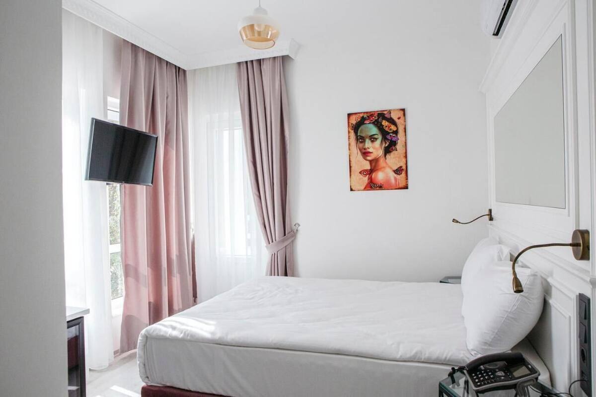 Quarto de hotel com uma cama, televisão na parede, um quadro e telefone na mesinha de cabeceira.