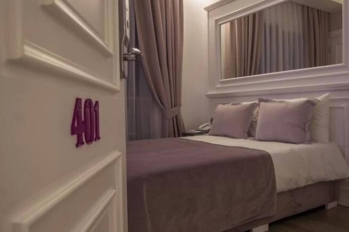 Quarto de hotel com uma cama, uma janela com cortinas e uma porta com o número.