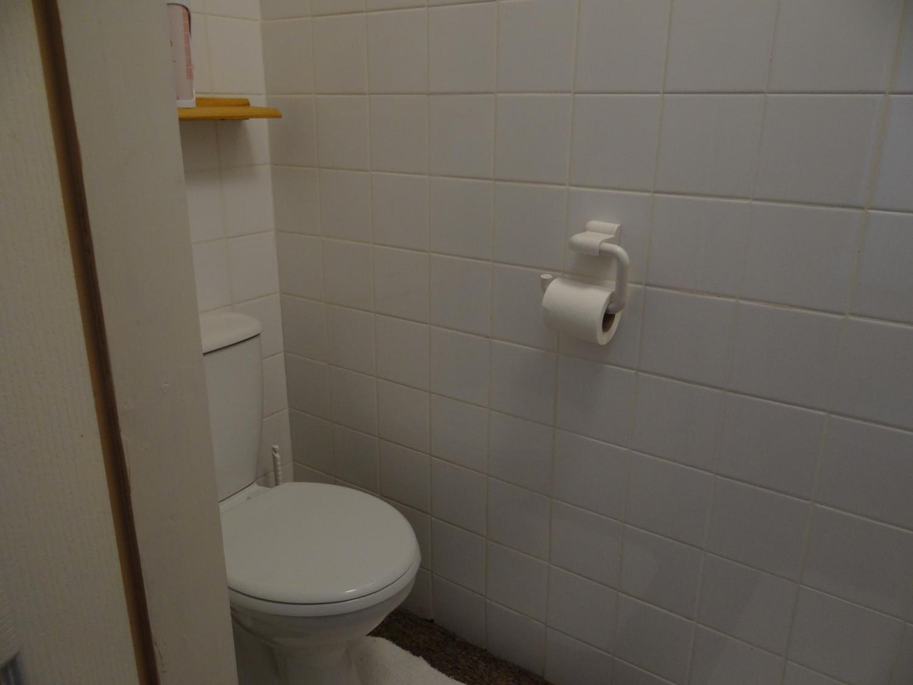 Casa de banho pequena e limpa, com sanita, uma toalha branca no chão e um porta-papel higiénico na parede.