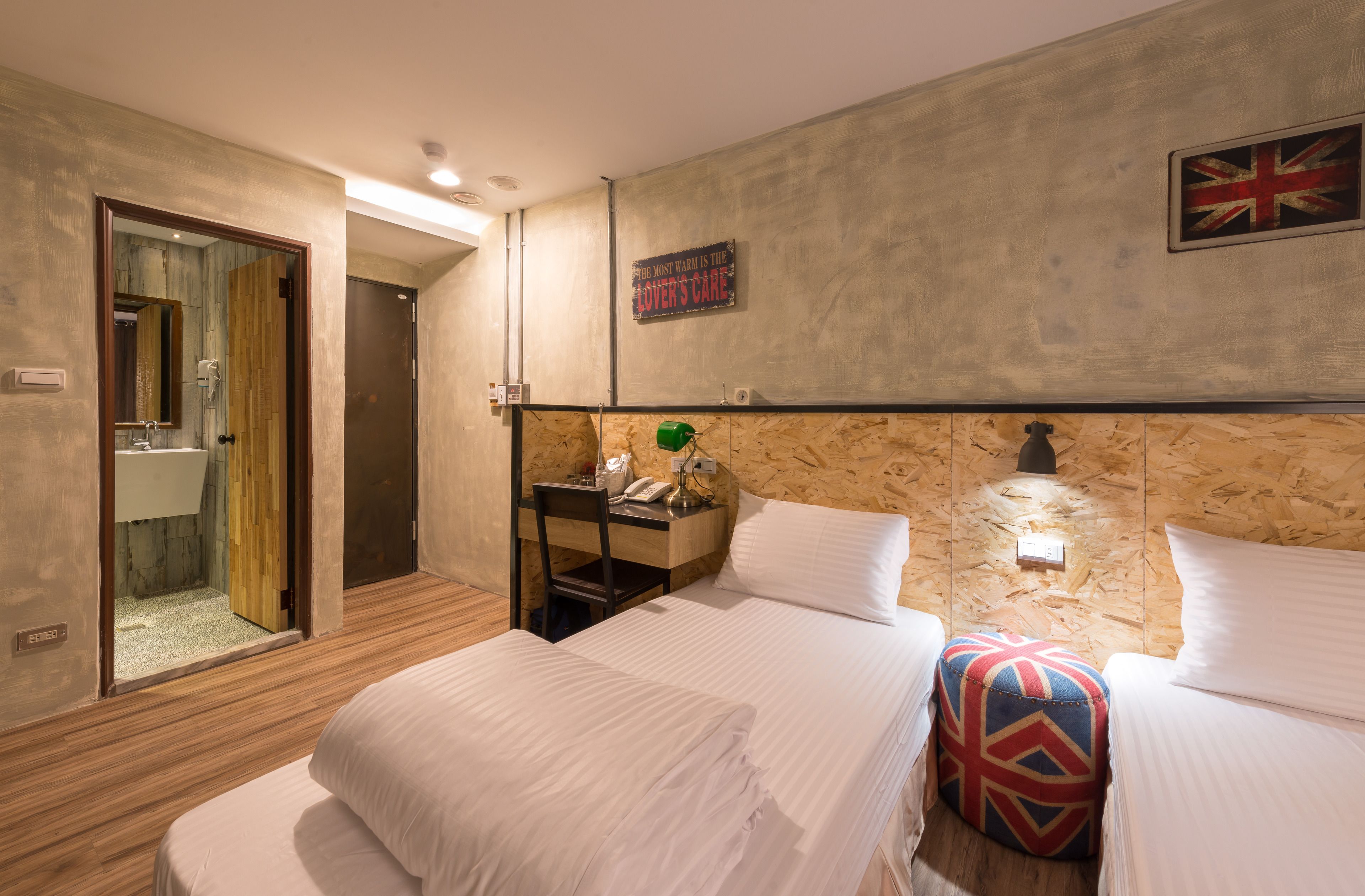 Quarto de hotel com piso de madeira, cama com roupa de cama branca, escrivaninha com telefone e luminária fixada na parede.