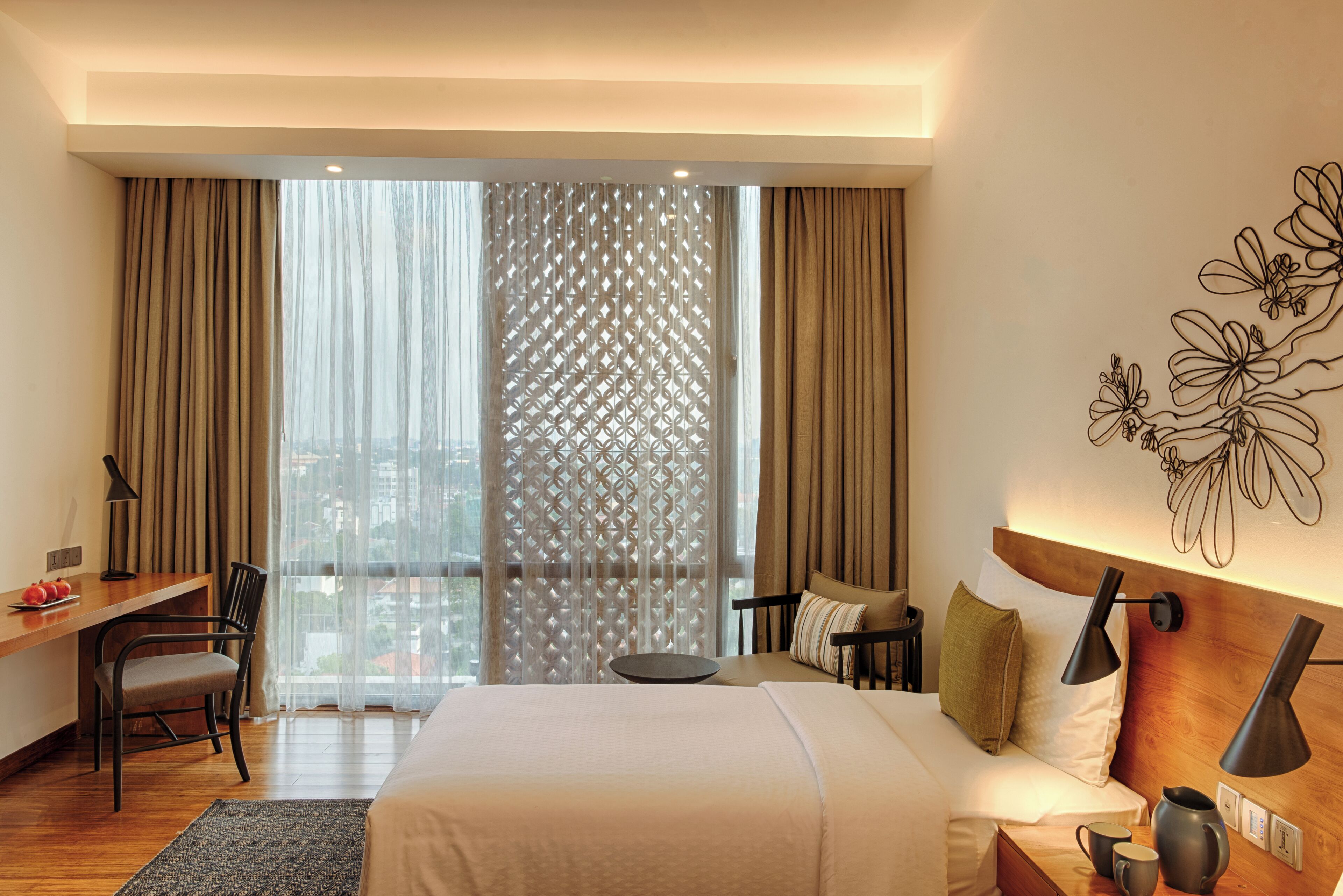 Quarto de hotel com cama, escrivaninha, cadeira e vista pela janela com cortinas.