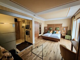 Quarto de hotel com chão de madeira, cama com várias almofadas, guarda-roupa de madeira, cabine de duche em vidro e uma pequena mesa com uma cadeira.