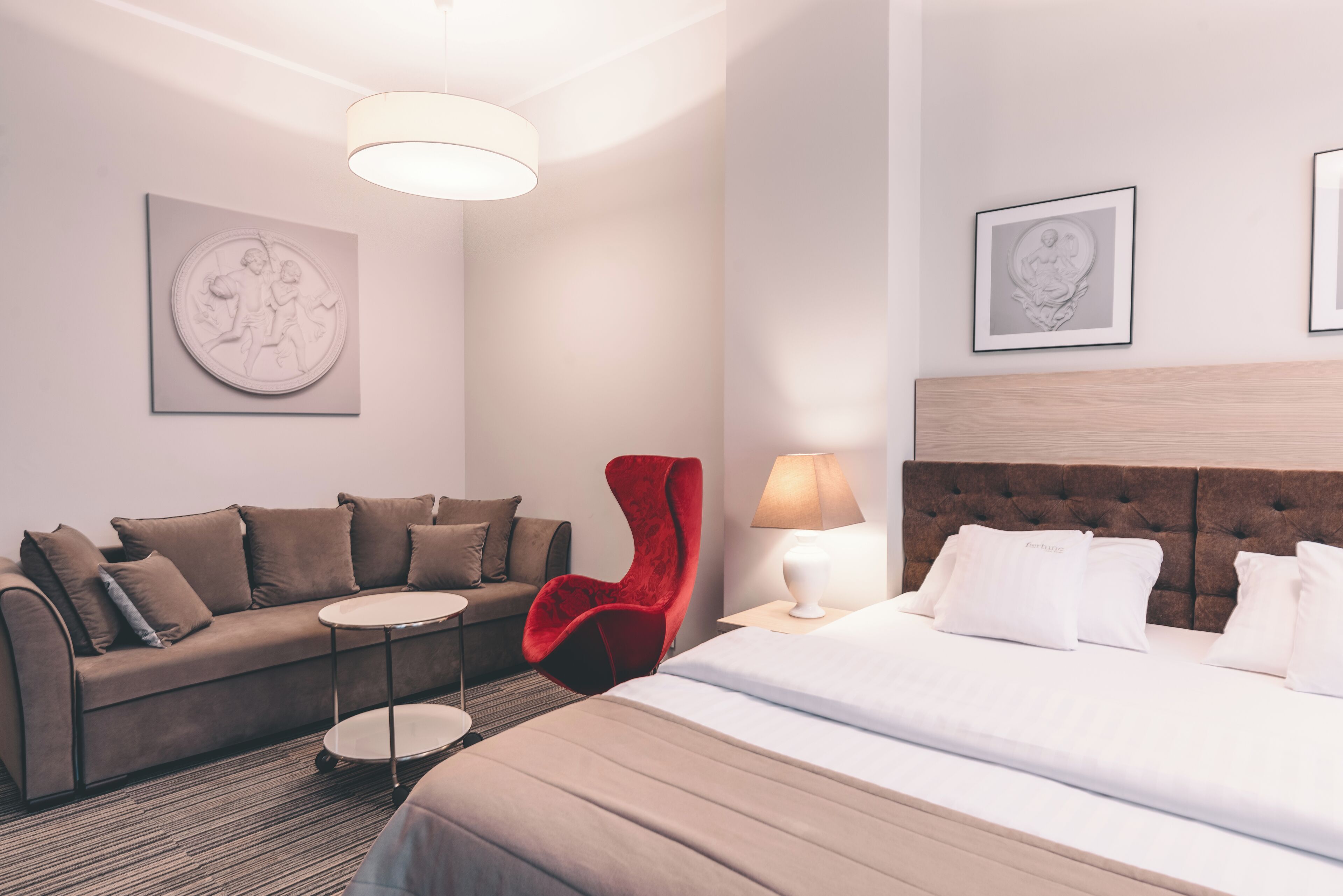 Quarto de hotel com uma cama, uma cadeira vermelha, uma mesinha, um sofá e duas obras de arte fixadas na parede.