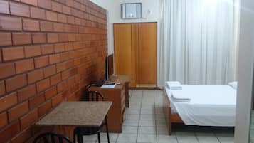 Quarto pequeno e aconchegante com parede de tijolos, uma escrivaninha de madeira com uma cadeira, cama com roupa de cama branca e um guarda-roupa de madeira.