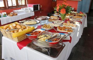 Uma mesa de buffet com diversos alimentos e utensílios, com uma parede vermelha, janelas e um arranjo de flores ao fundo.