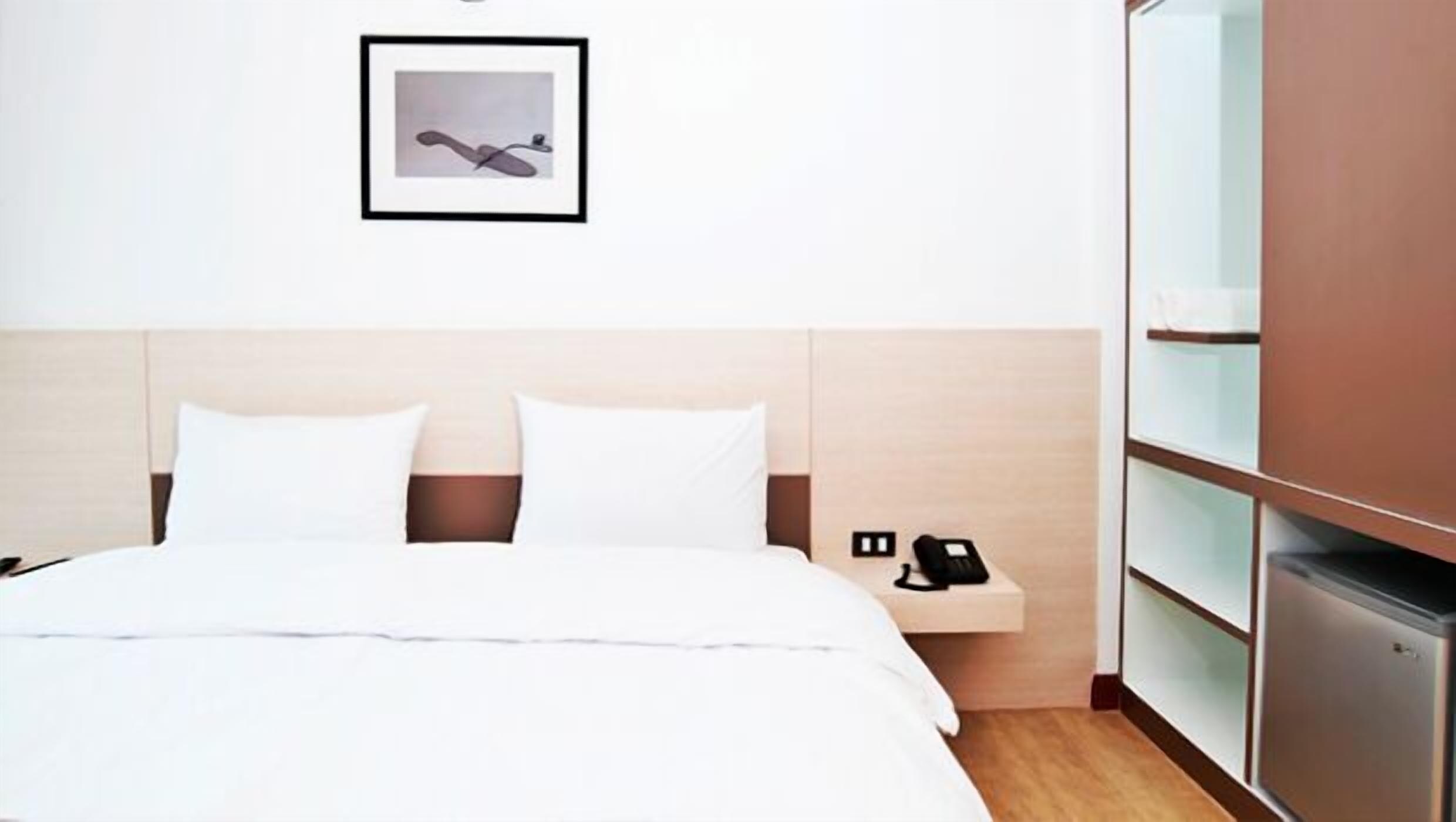 Quarto de hotel com uma cama, almofadas brancas, mesa de cabeceira com telefone e um quadro na parede.