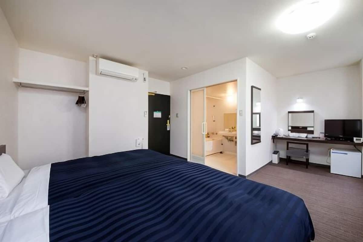Quarto de hotel com cama, escrivaninha, televisão e banheiro visível através de uma porta aberta.