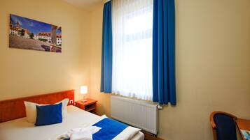 Quarto de hotel com cama, mesa de cabeceira, luminária, janela com cortinas azuis e um quadro na parede.