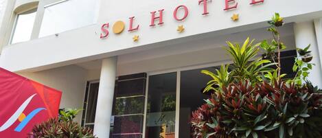 Exterior do Hotel Sol com uma placa grande, janelas amplas e plantas em vasos na frente.