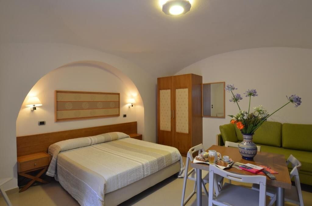 Quarto de hotel com cama, escrivaninha com cadeira, sofá verde, vaso com flores e espelho fixado na parede.