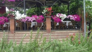 Uma área de estar coberta ao ar livre, com mesas e cadeiras, rodeada por plantas e flores em vasos.