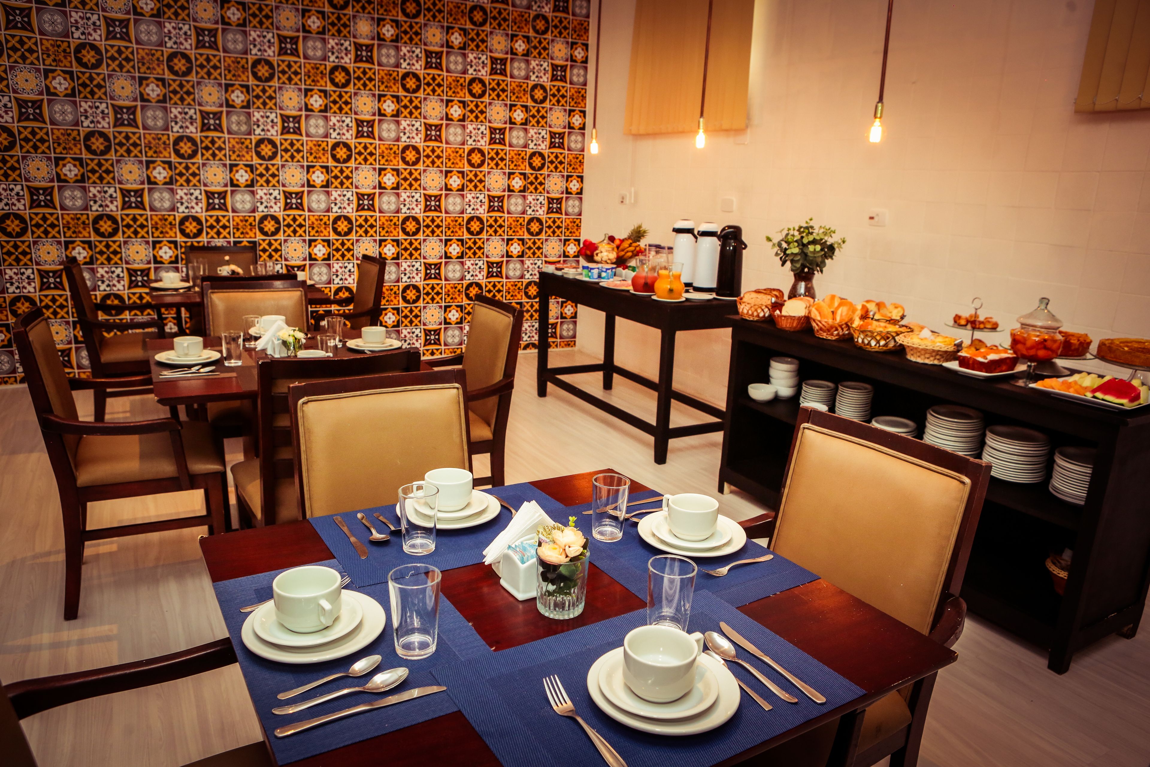 Espaço de refeições com parede estampada, buffet com diversos alimentos e mesas organizadas com cadeiras.