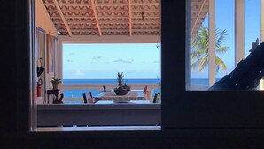 Vista de um quarto com janela para a praia, apresentando um teto com padrão geométrico, uma mesa com planta em vaso e vista do oceano.