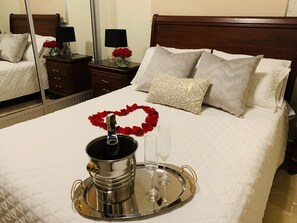 Uma cama bem arrumada com um arranjo de rosas vermelhas em formato de coração, um balde de champanhe e duas taças de vinho sobre uma bandeja.
