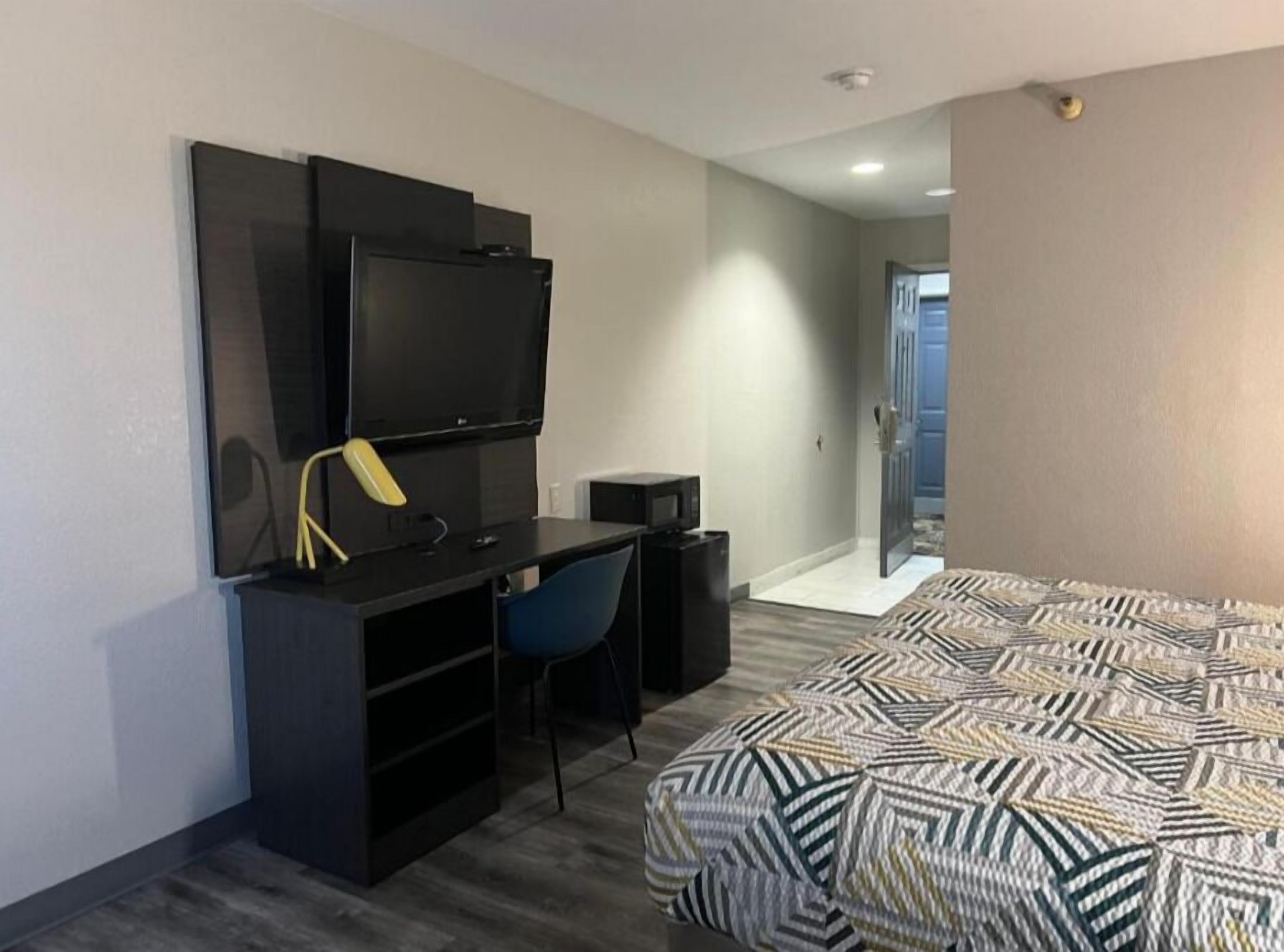 Quarto de hotel com cama, escrivaninha com televisão, cadeira e mesa pequena.
