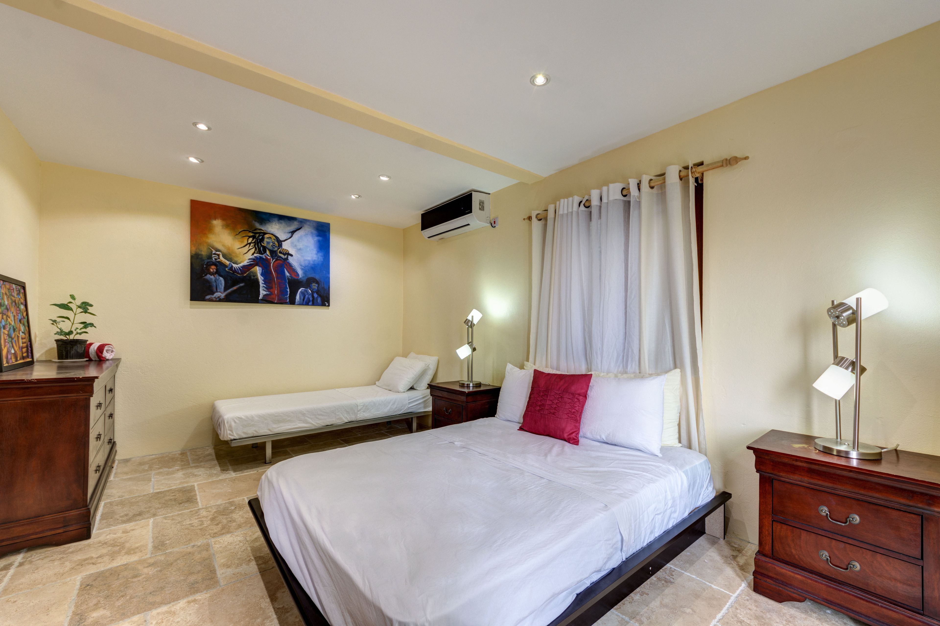 Quarto com uma cama grande, uma cama menor, mesa de cabeceira com abajur, cômoda e um quadro na parede.