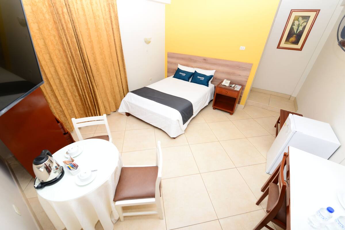 Quarto de hotel com cama, escrivaninha, cadeira, mesa, televisão e um quadro na parede.