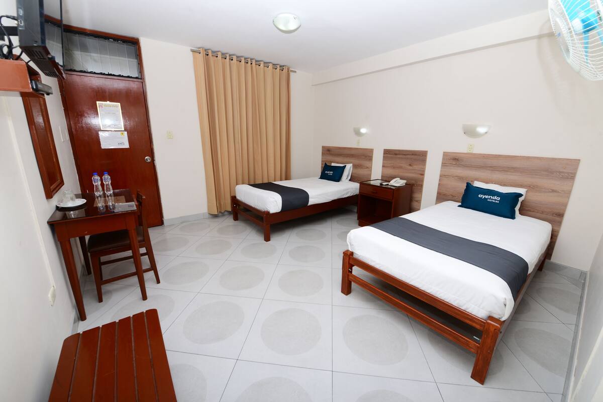 Quarto de hotel com duas camas de solteiro, uma escrivaninha de madeira, uma cadeira, uma mesa pequena com garrafas e uma cortina.