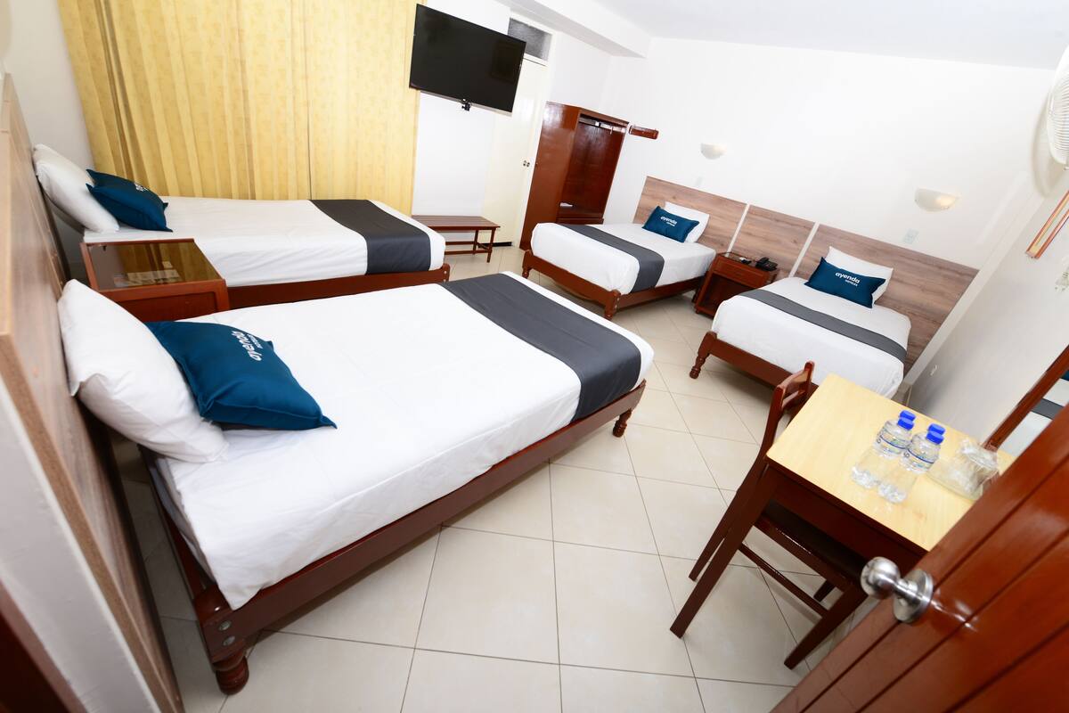 Quarto de hotel com quatro camas de solteiro, uma escrivaninha de madeira, uma cadeira, uma TV fixada na parede e uma porta com puxador.