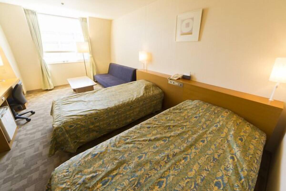 Quarto de hotel com duas camas de solteiro, uma escrivaninha, uma cadeira e uma janela com cortinas.