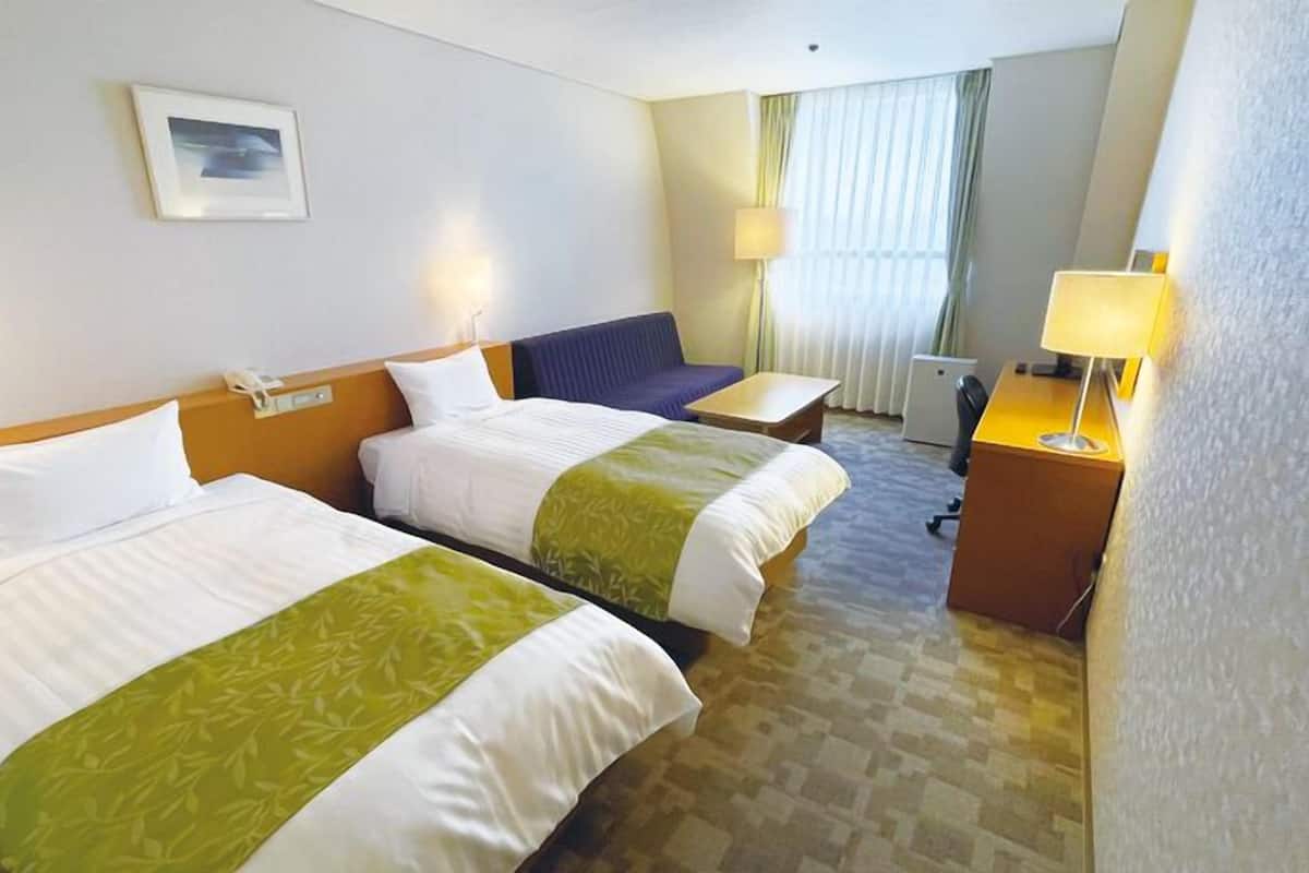 Quarto de hotel com duas camas, uma escrivaninha, uma cadeira, um abajur e uma janela com cortinas.