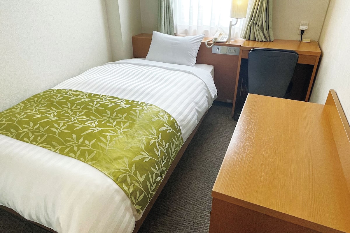 Quarto de hotel com cama, escrivaninha, cadeira, abajur e telefone.