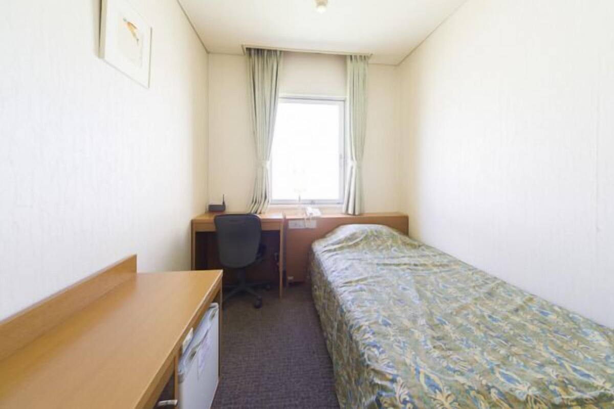 Quarto pequeno com uma cama de solteiro, escrivaninha e cadeira, janela com cortinas e radiador.