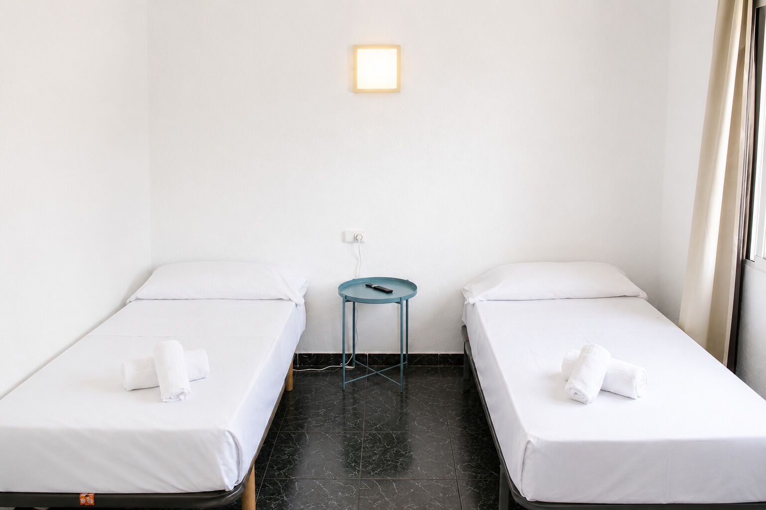 Quarto com duas camas de solteiro, uma mesinha, uma tomada e janela com cortinas.