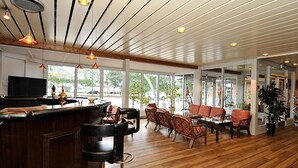 Ampla área de bar com piso de madeira, banquetas de couro e iluminação pendente. Há uma área de estar com cadeiras de vime e uma mesa, e grandes janelas que proporcionam iluminação natural.