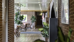 Um corredor com parede de tijolos, piso de madeira e plantas em vasos. Há uma área de estar com cadeiras e uma mesa ao fundo.