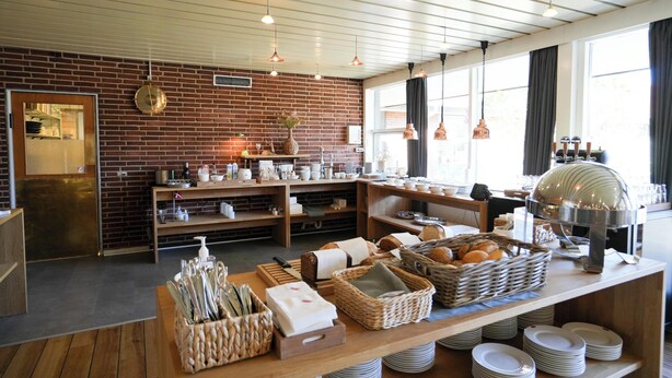 Buffet de café da manhã com diversos alimentos e utensílios, um espaço para café e uma parede de tijolos ao fundo.