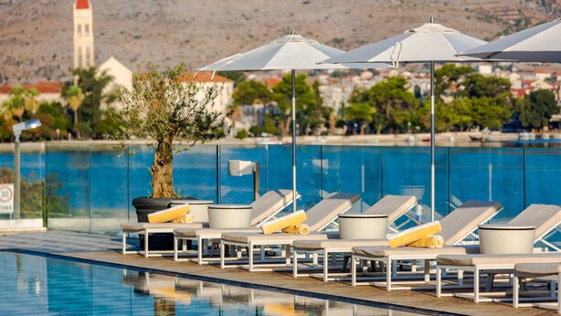 Área junto à piscina com espreguiçadeiras e guarda-sóis, uma árvore e um edifício com telhado vermelho ao fundo.