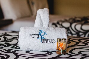 Toalha branca com o logotipo "HOTEL FLAMINGO", garrafa de vidro com líquido laranja e uma colcha estampada.