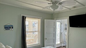 Quarto com cama, ventoinha no teto, janela com persianas, porta e TV fixada na parede.