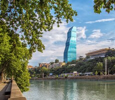 Um edifício moderno com um design curvo distinto, um rio ao primeiro plano e uma ponte com árvores à esquerda.