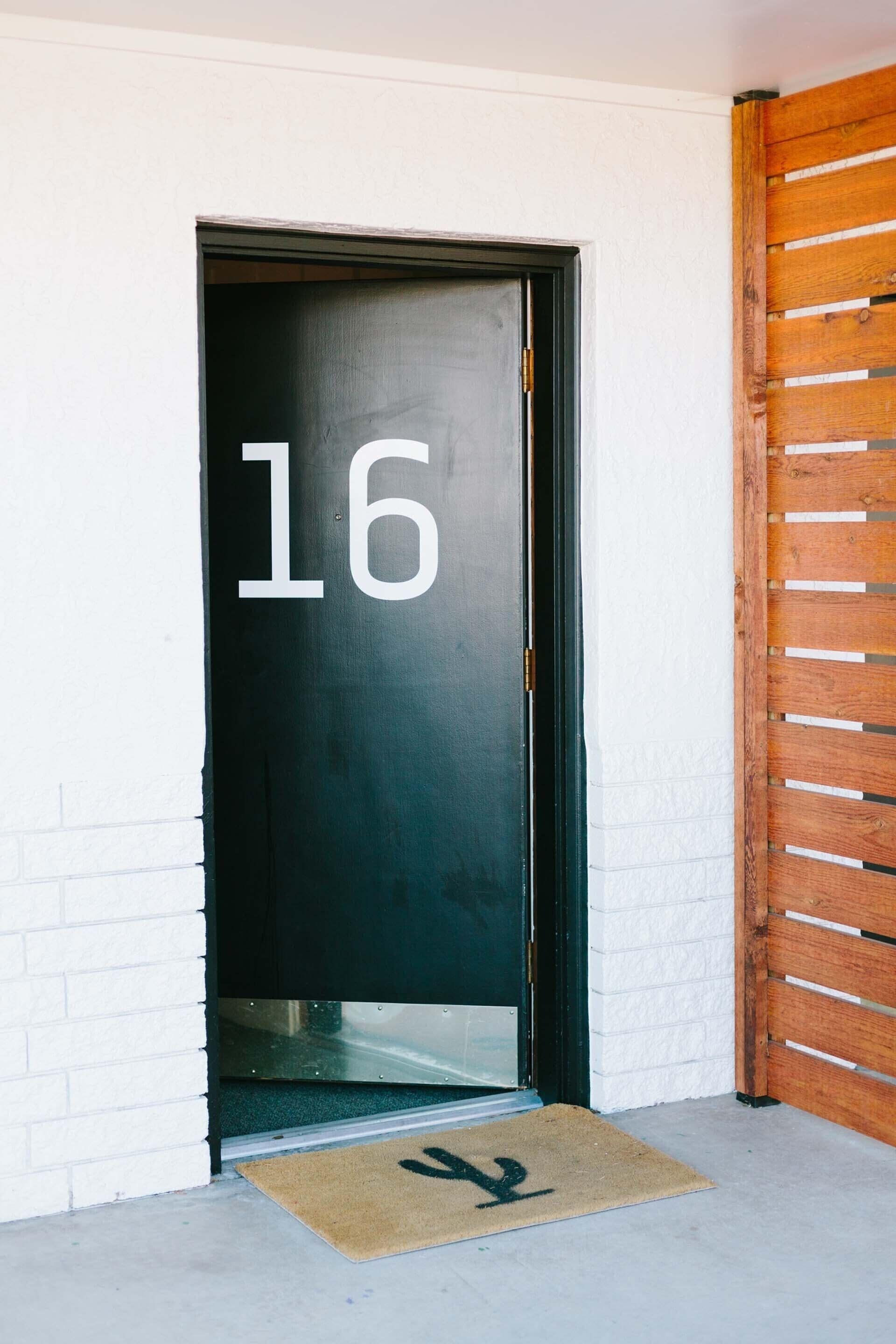 Uma porta preta com o número 16, uma parede de tijolos brancos e um vão de porta de madeira ripas.