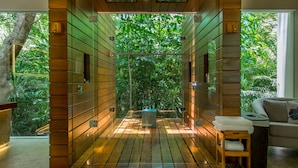 Quarto moderno com piso de madeira, divisória de vidro e vista para vegetação exuberante.