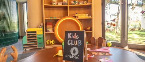 Uma mesa de madeira com uma placa que diz "Kids Club", rodeada por cadeiras e brinquedos infantis.