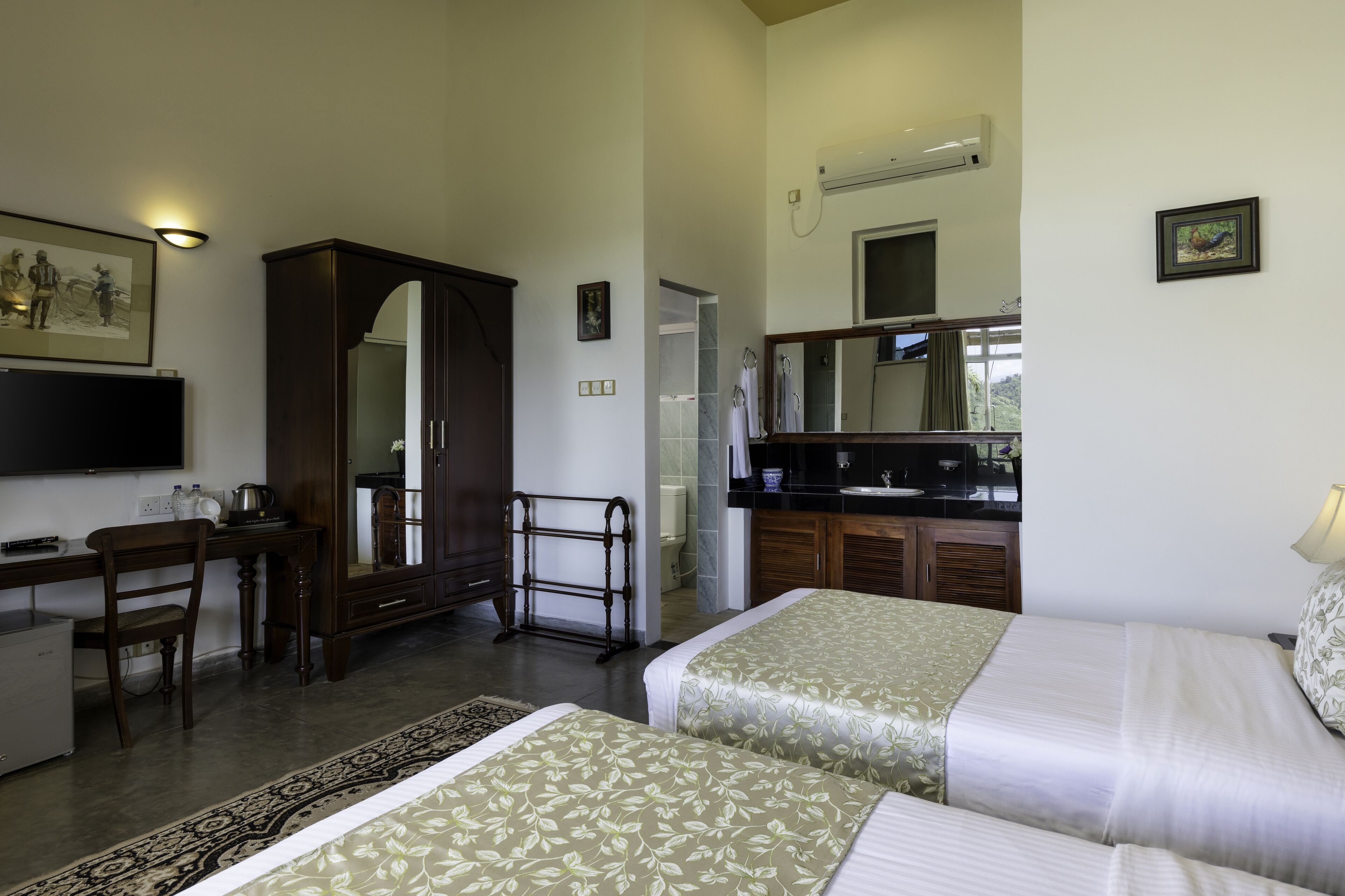 Quarto de hotel com duas camas, uma escrivaninha, um guarda-roupa, um espelho e um banheiro visível através de uma porta aberta.