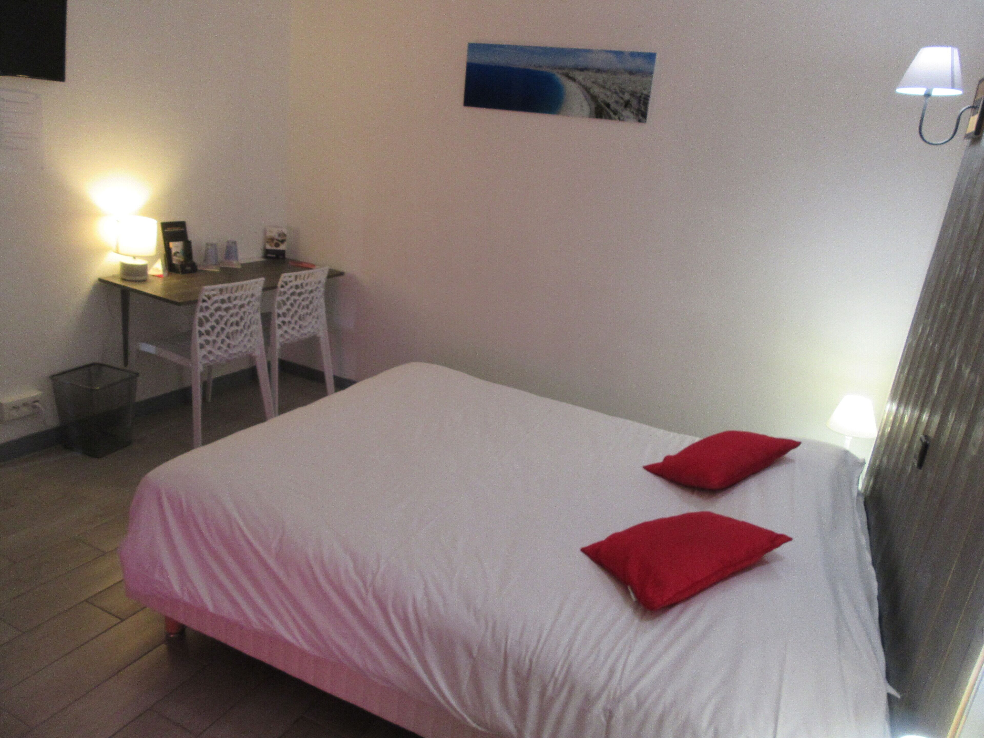 Quarto de hotel com cama, escrivaninha, duas cadeiras, lixeira e quadro fixado na parede.