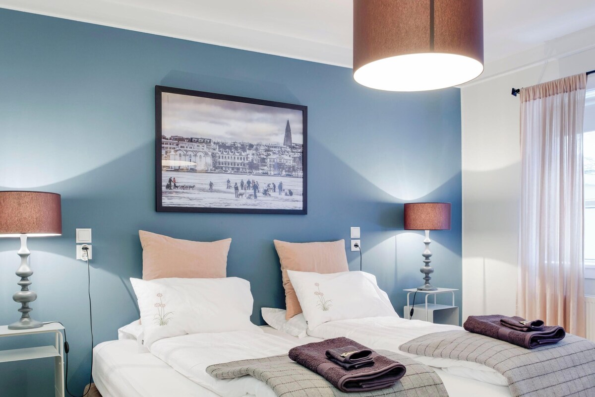Quarto de hotel com cama, duas mesinhas de cabeceira, abajures, um quadro pendurado na parede e uma janela com cortinas.
