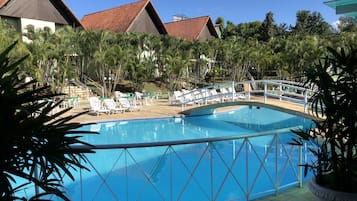 Piscina com cerca geométrica, rodeada por espreguiçadeiras e palmeiras, com edifícios ao fundo.