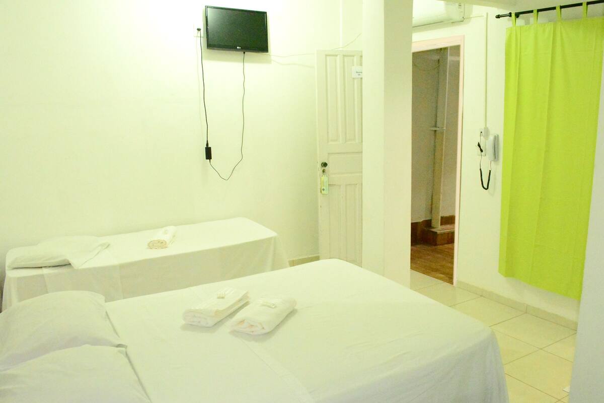 Quarto de hotel com uma cama, uma mesinha com telefone e uma televisão na parede.