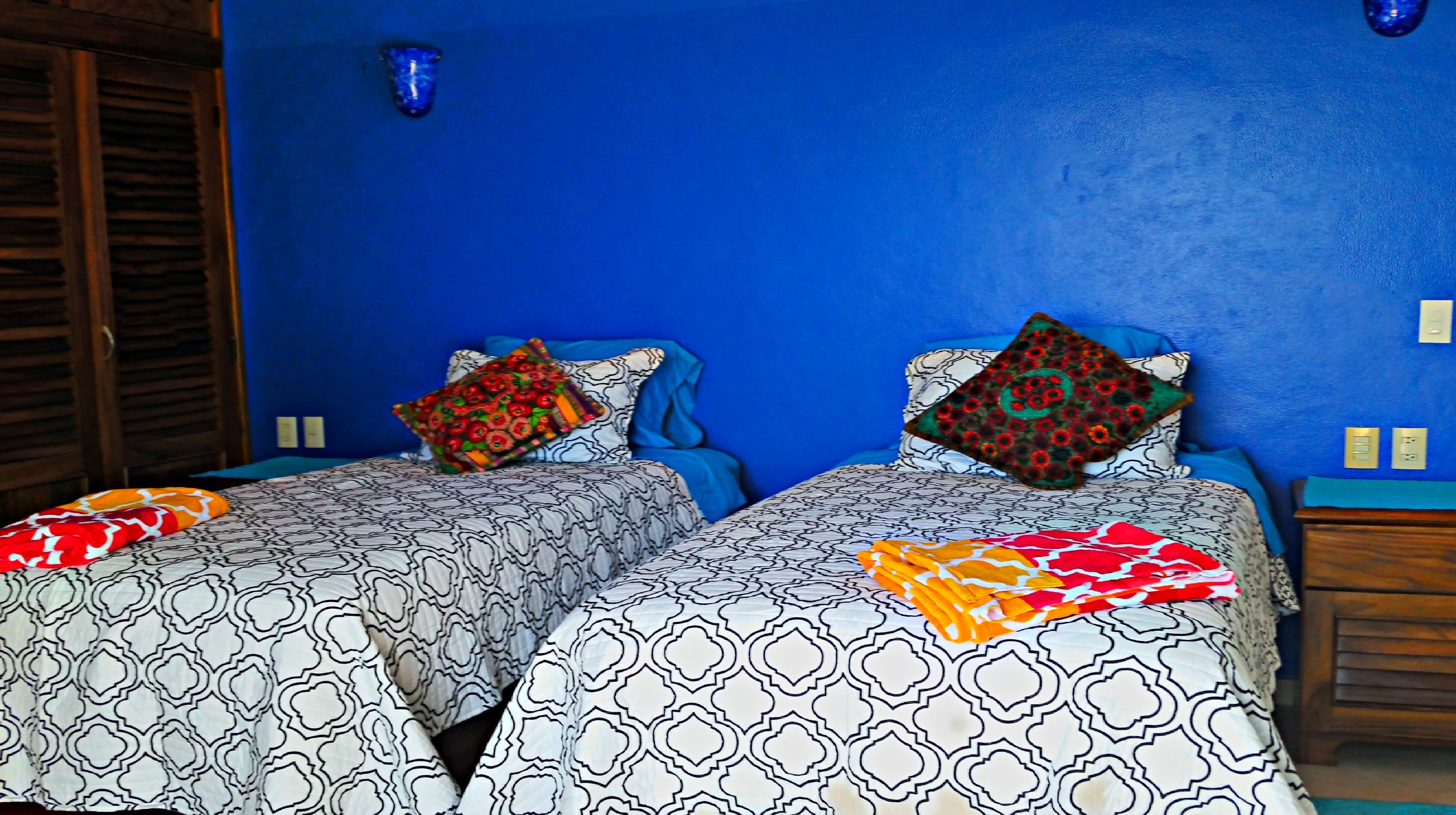 Quarto com duas camas, cada uma com roupa de cama e almofadas estampadas, parede azul e mesa de cabeceira de madeira com abajur.