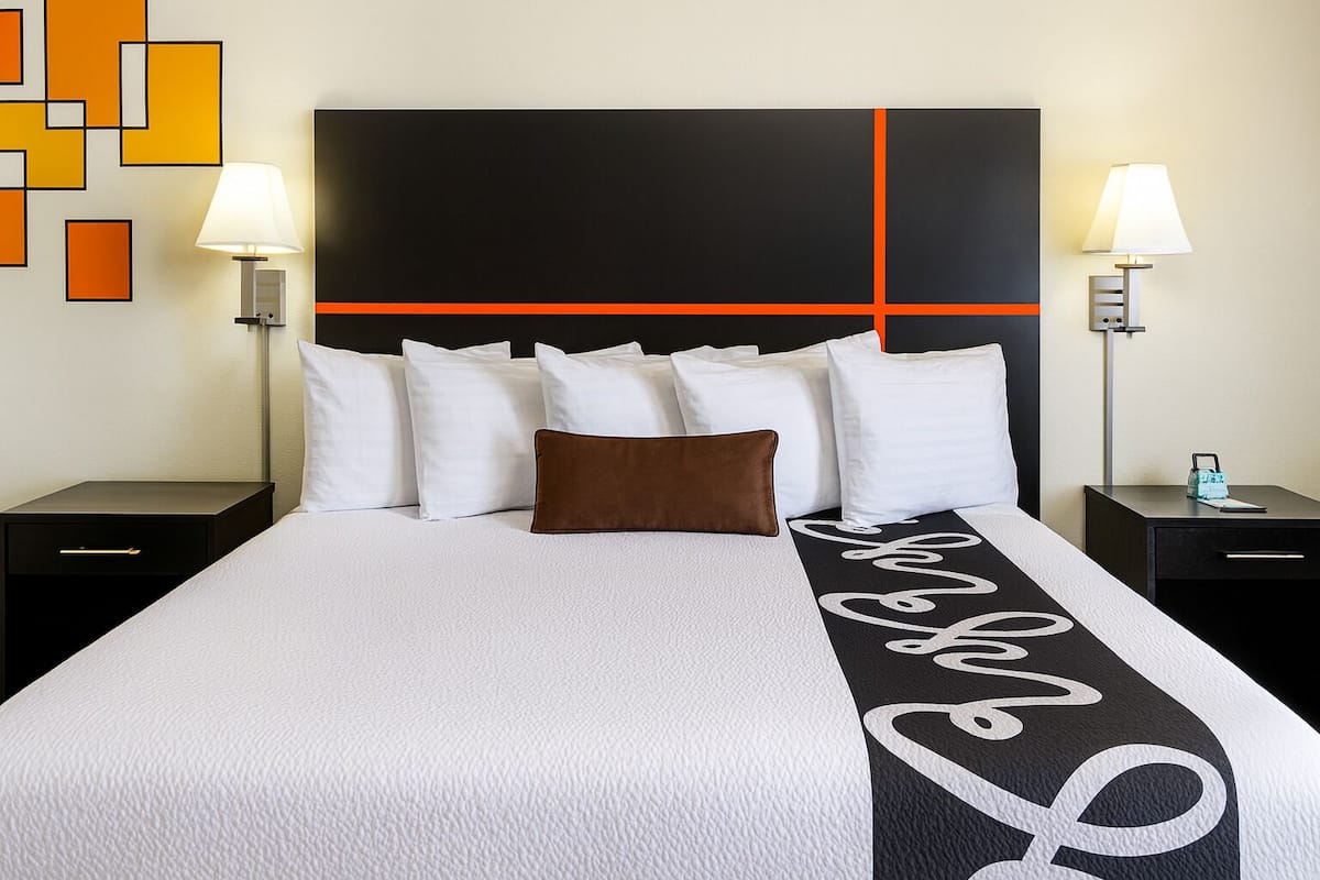 Quarto de hotel com cama grande, roupa de cama branca, cabeceira preta e laranja, duas mesas de cabeceira com abajures e um objeto decorativo na parede.
